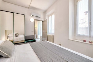 Quarto de hotel com cama, mesa de cabeceira, espelho, radiador e janela com cortinas.
