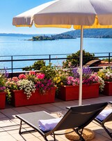 Um terraço com vista para um lago, vasos de flores vermelhas e duas cadeiras de descanso.