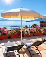 Terraço com duas poltronas de descanso, um grande guarda-sol e vasos de flores, com vista para um lago e montanhas.