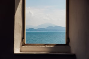 Janela com vista para um mar tranquilo e montanhas ao longe.