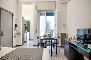 Quarto de hotel com cama, mesa de jantar com cadeiras, pequena cozinha e televisão sobre um suporte.