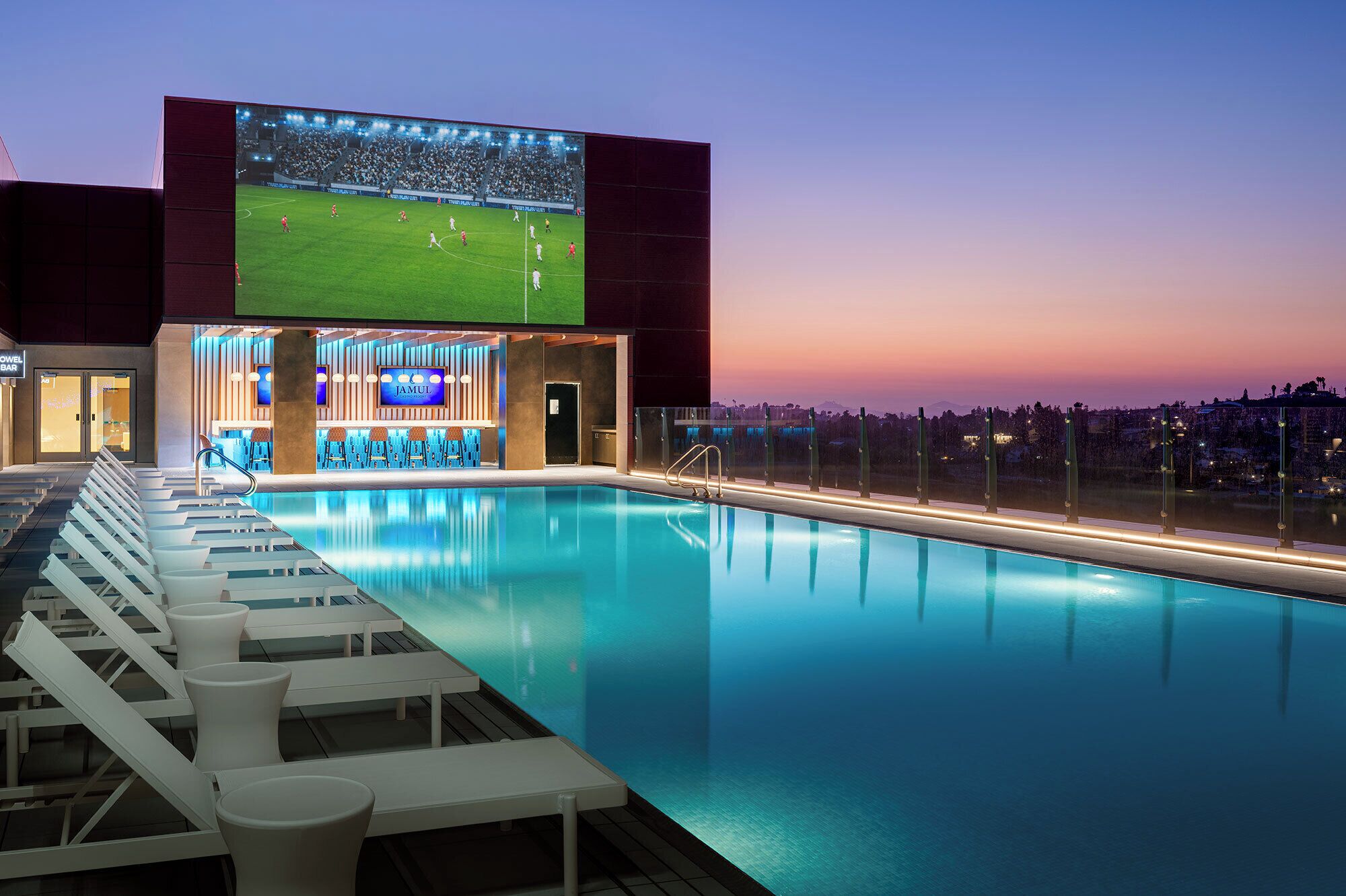 Área de piscina com espreguiçadeiras, bar e uma grande tela exibindo uma partida de futebol.