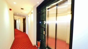 Corredor de hotel com tapete vermelho, elevador de vidro e uma porta no final.