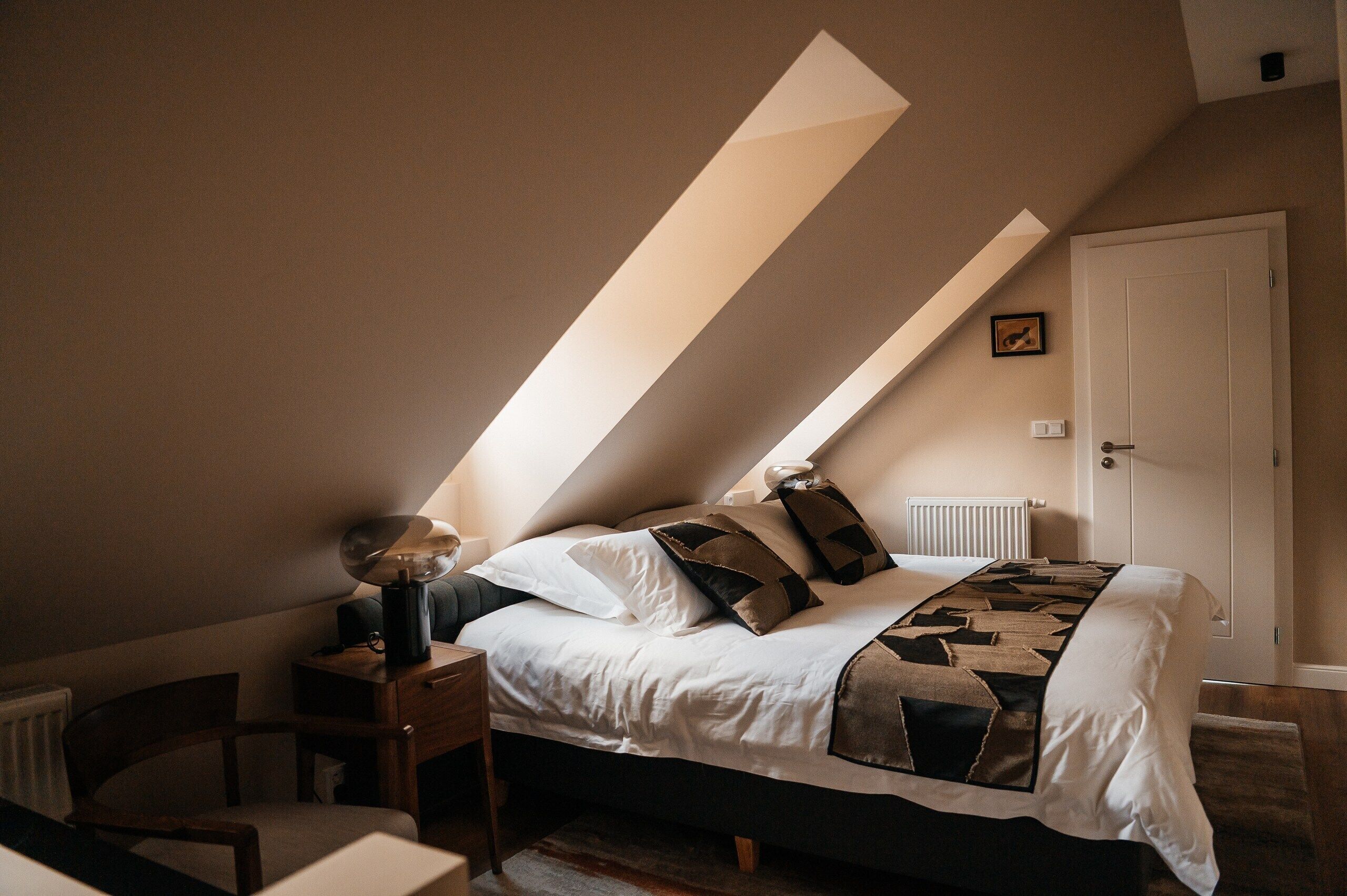 Um quarto com teto inclinado, uma cama com roupa de cama branca e manta estampada, uma mesa de cabeceira com abajur e um radiador.