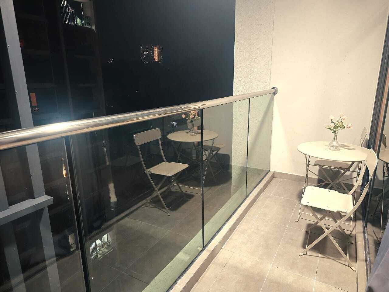 Uma varanda com guarda-corpo de vidro, uma mesa redonda com cadeiras e uma pequena mesa com um vaso.