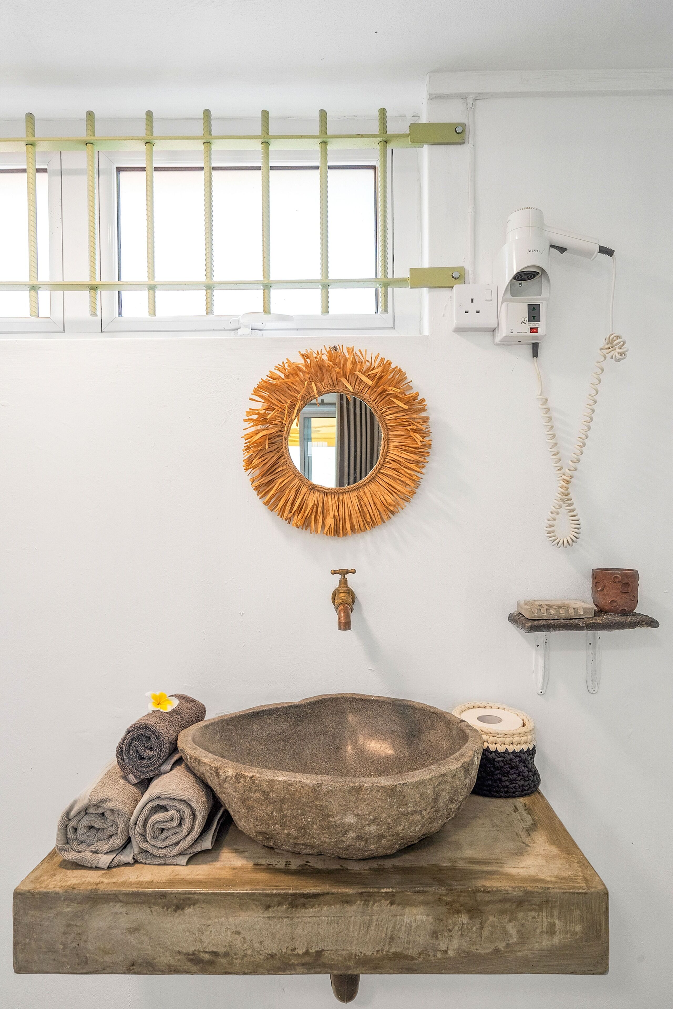 Casa de banho com lavatório de pedra, porta-toalhas, espelho com padrão de franjas, secador de cabelo e pequena prateleira com produtos de higiene pessoal.