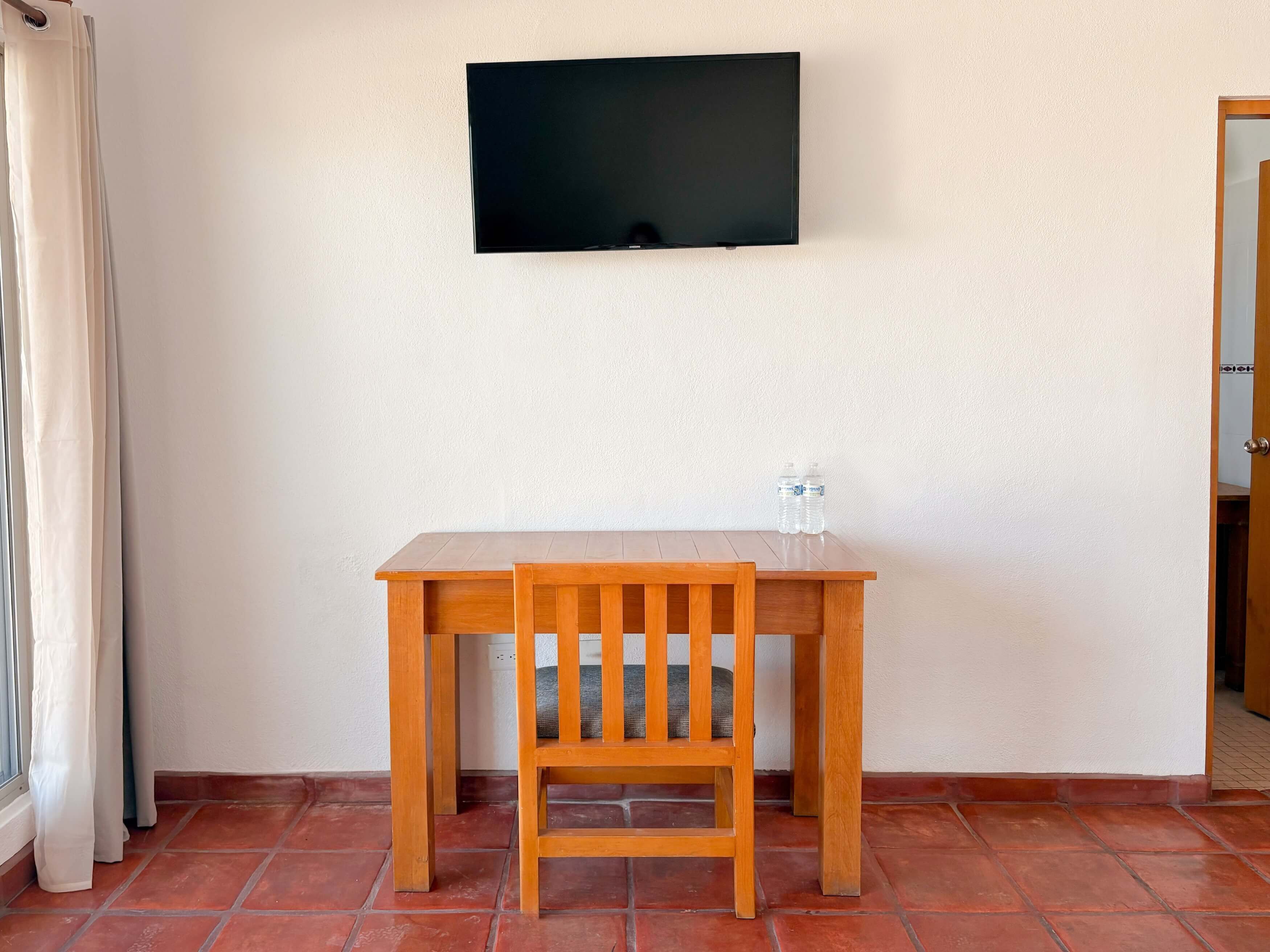 Um quarto com uma mesa de madeira, uma cadeira, uma TV fixada na parede e uma garrafa de água sobre a mesa.