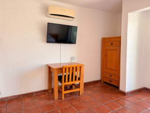 Um quarto pequeno com uma mesa de madeira, uma televisão na parede, um armário de madeira e piso de azulejos.