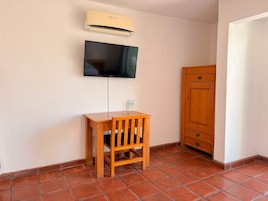 Um quarto pequeno com uma mesa de madeira, uma televisão na parede, um armário de madeira e piso de azulejos.