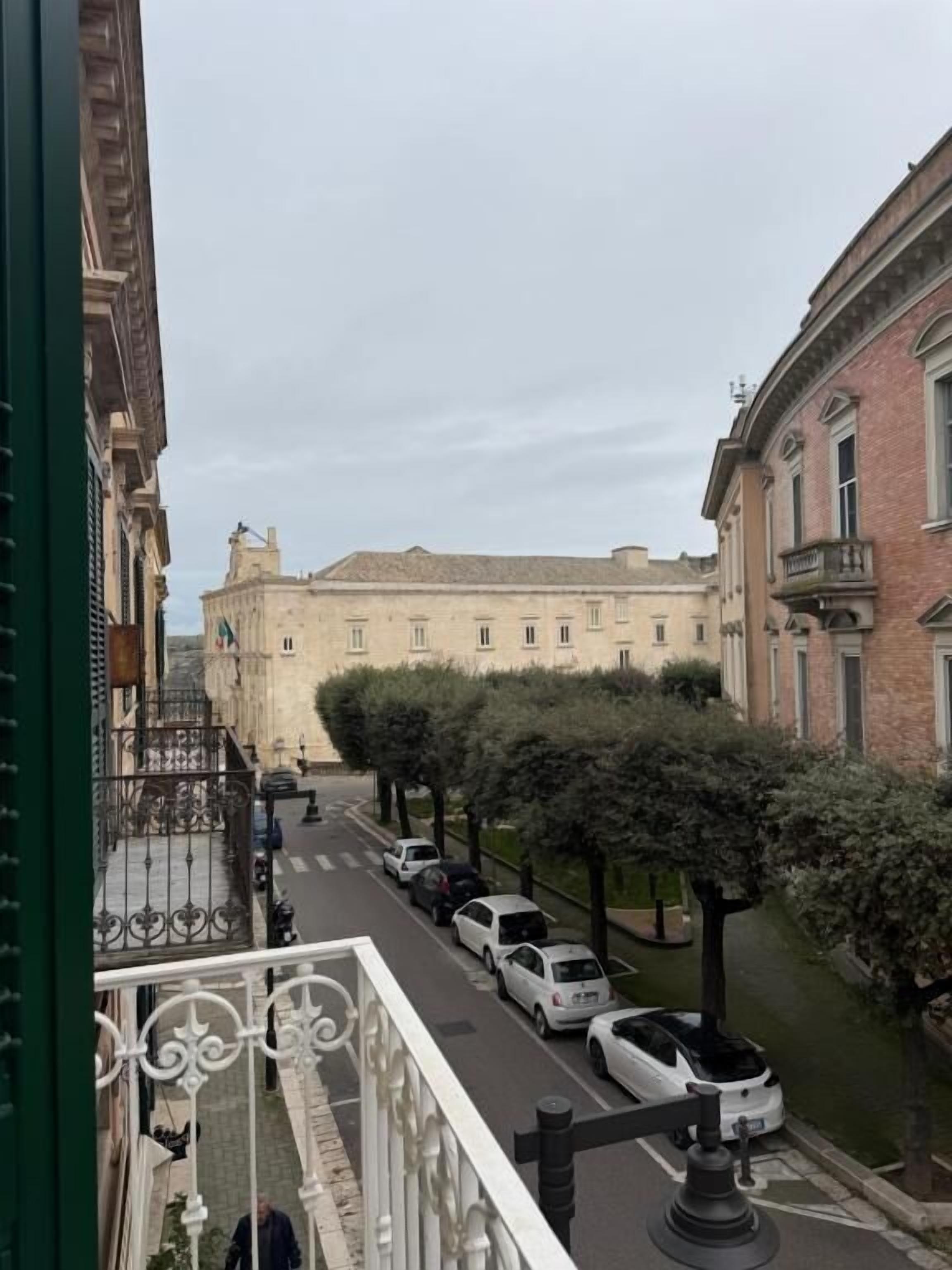 Vista da rua com um edifício, carros estacionados e uma calçada arborizada.