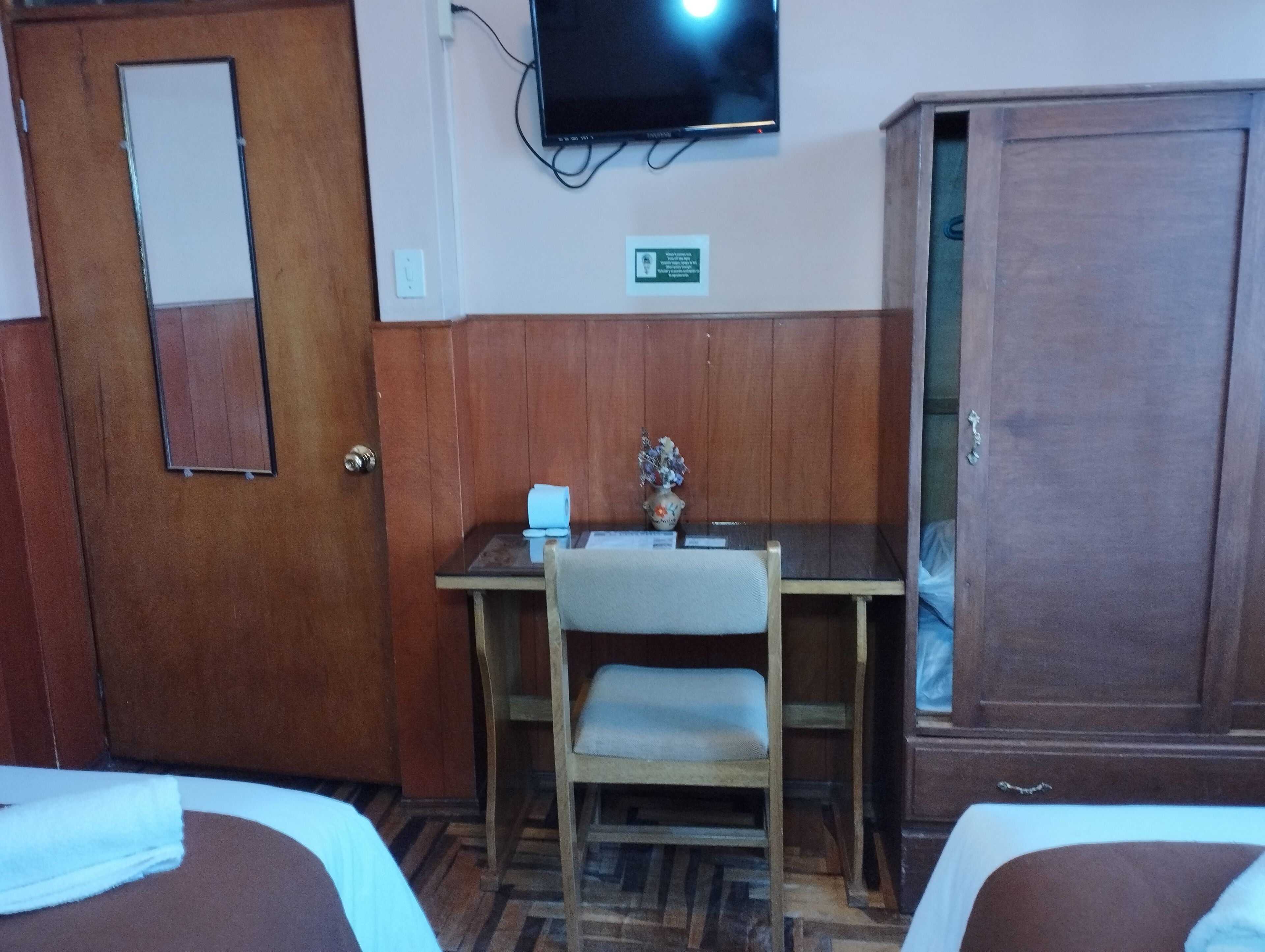 Quarto com escrivaninha de madeira, uma cadeira, televisão na parede e guarda-roupa.