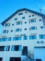 Um hotel com vários andares, fachada branca, várias janelas e uma escadaria visível que leva à entrada.