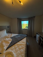 Quarto de hotel com cama, escrivaninha, janela com cortinas e vista para o verde lá fora.