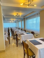Uma sala de banquetes com filas de mesas e cadeiras, janelas amplas e um lustre moderno.