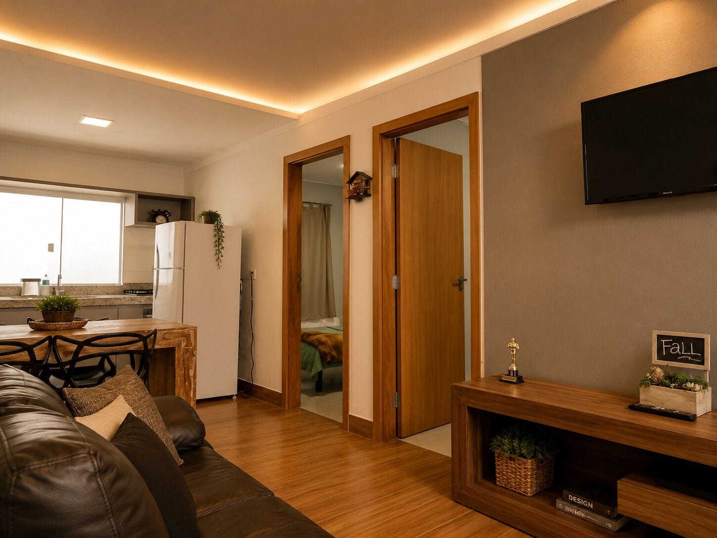 Uma sala de estar moderna com sofá de couro, piso de madeira, televisão fixada na parede e uma cozinha ao fundo.