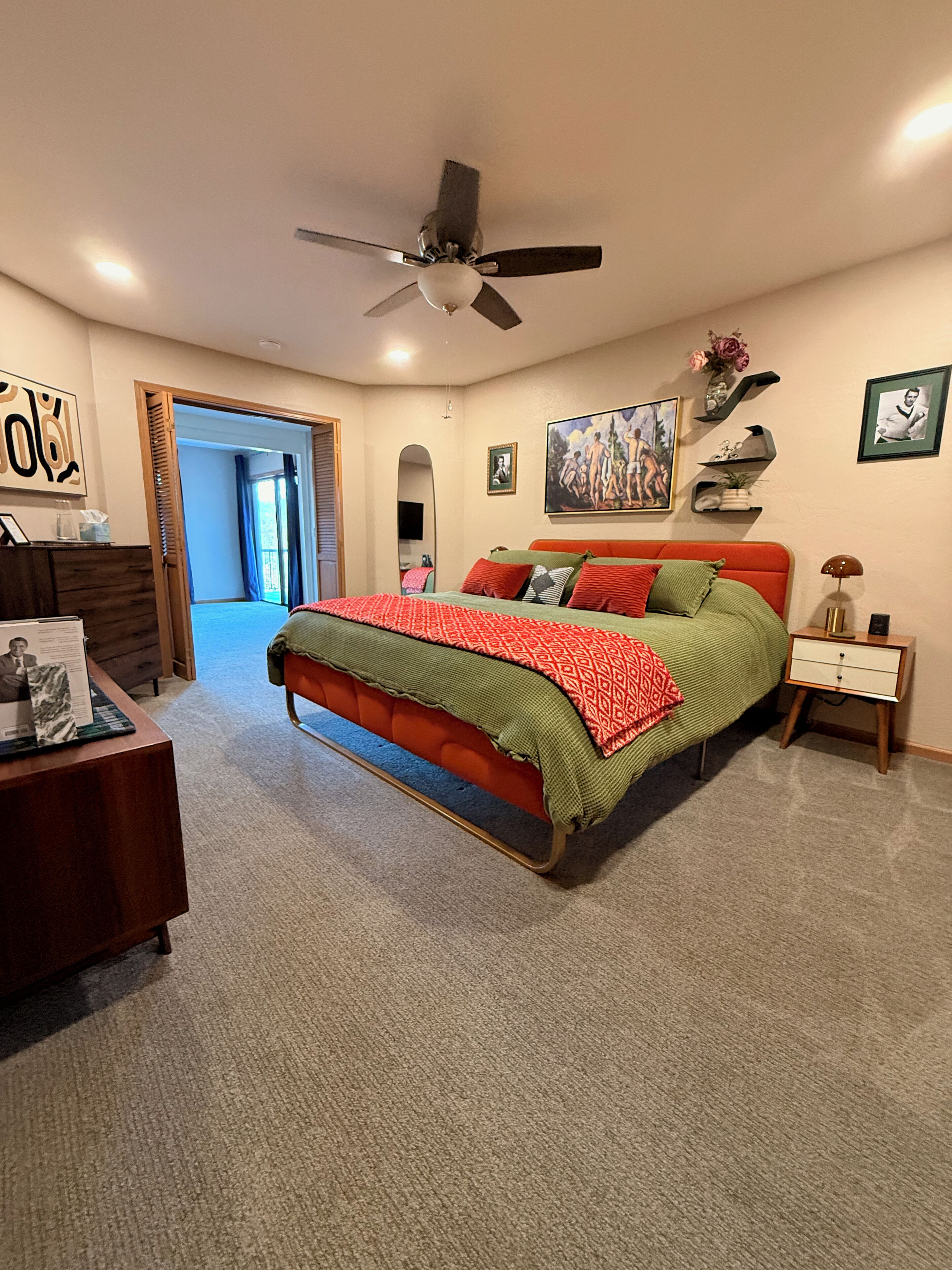 Quarto com cama, mesa de cabeceira, cômoda, ventilador de teto e porta que dá para outro espaço.