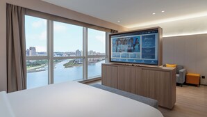 Quarto de hotel com uma televisão grande em um armário de madeira, uma cama, vista para um rio e para a paisagem urbana, e um banquinho laranja pequeno.