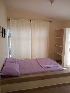 Um quarto com cama, janela com cortinas, prateleira na parede e ventilador de teto.