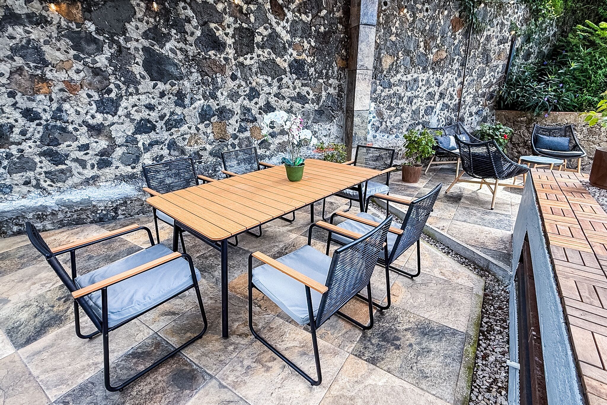 Um pátio com uma mesa e cadeiras de madeira, cercado por um muro de pedra e plantas em vasos.