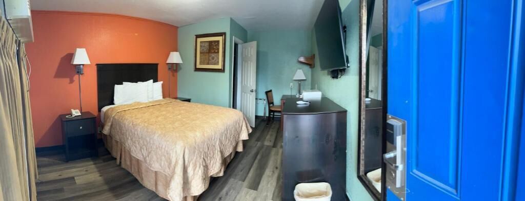 Quarto de hotel com cama, mesas de cabeceira, escrivaninha, cadeira, televisão e porta.