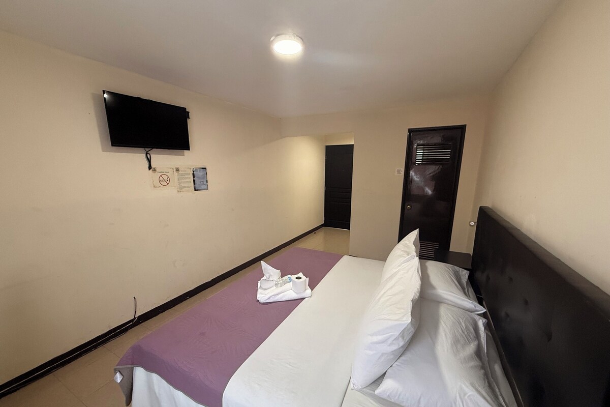 Quarto de hotel com uma cama, televisão fixada na parede e porta que dá para outro espaço.