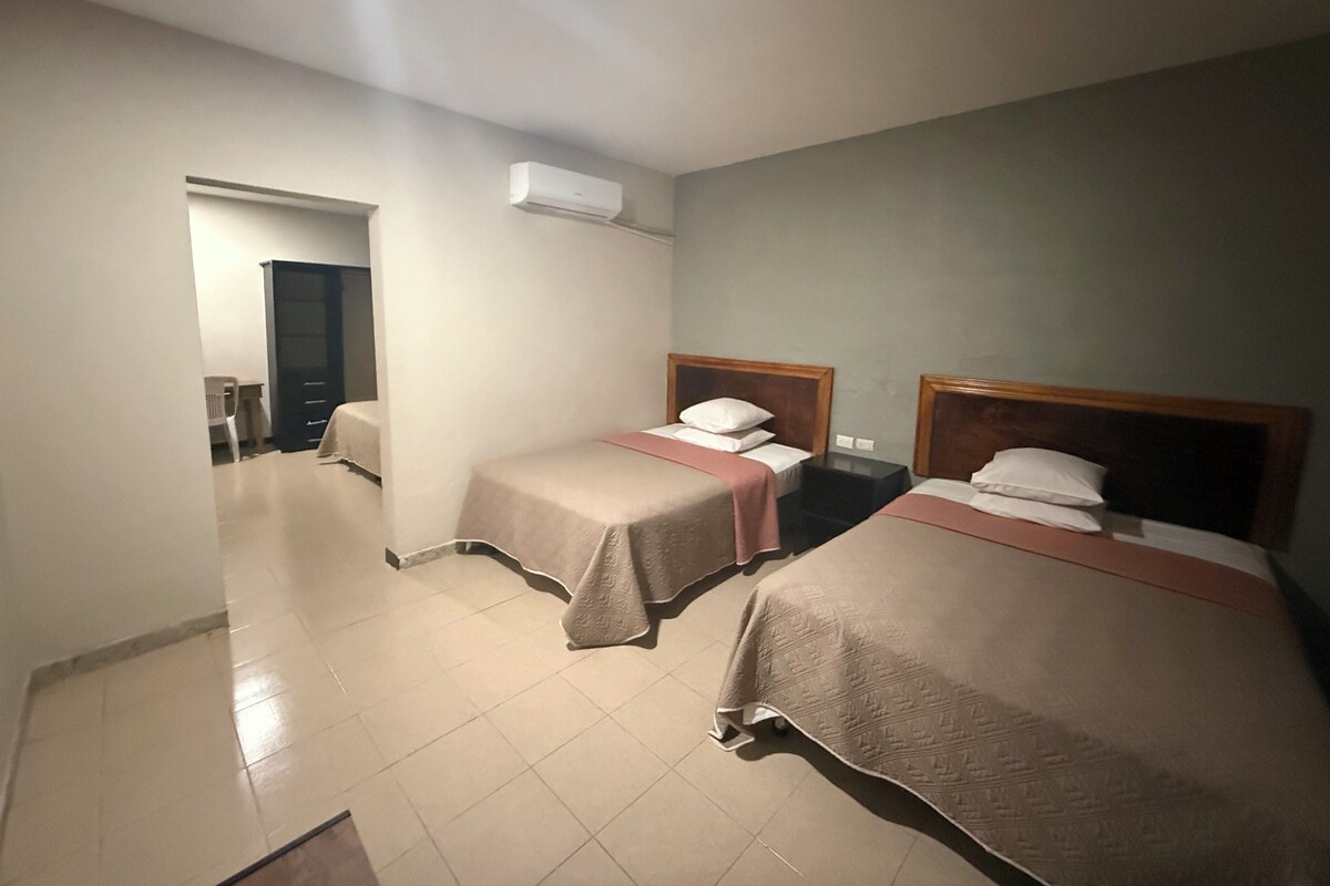 Quarto de hotel com três camas, uma escrivaninha e ar condicionado.