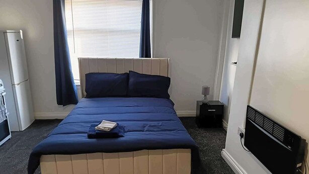 Cama bem arrumada com cobertura azul, pequena mesa de cabeceira com abajur, aquecedor e janela com persianas.
