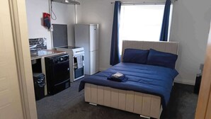Quarto de hotel pequeno e funcional, com uma cama, cozinha compacta, frigorífico, fogão e janela com cortinas.