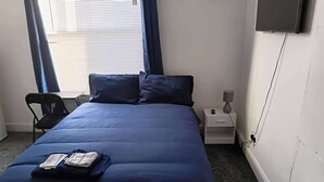 Quarto de hotel com cama, uma cadeira, mesa de cabeceira, luminária e televisão fixada na parede.