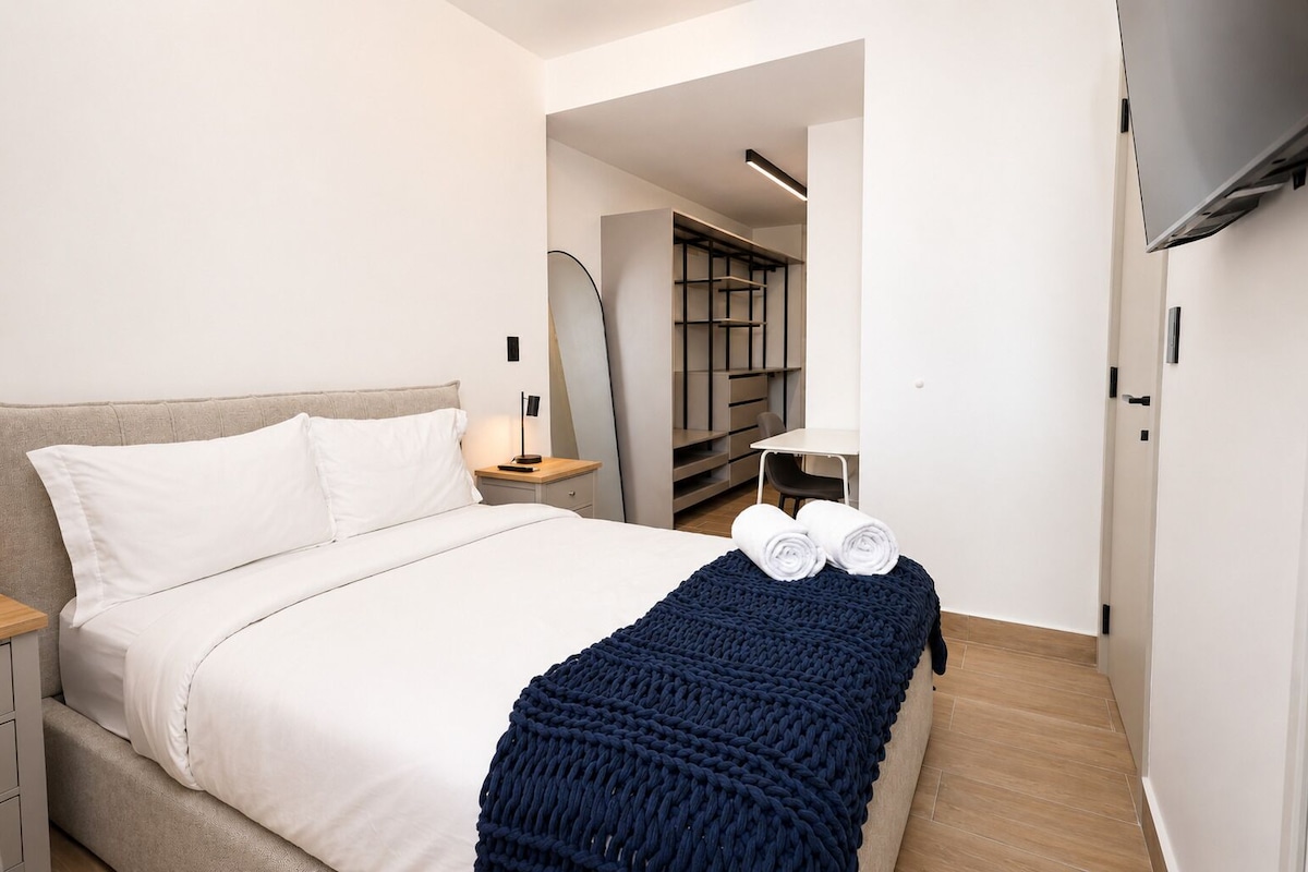 Quarto de hotel com uma cama, uma mesa de cabeceira, um abajur, uma toalha enrolada e um cobertor azul-escuro de tricô.