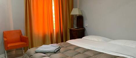 Quarto de hotel com cama, uma cadeira, abajur e janela com cortinas.