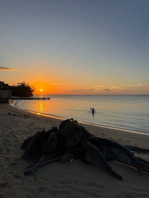 Cena de praia com um pôr do sol, mar calmo e uma grande formação rochosa em primeiro plano.