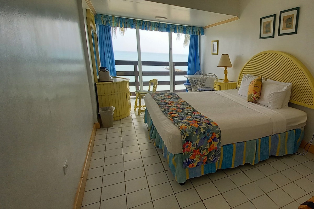 Quarto de hotel com cama, duas luminárias de cabeceira, janela com cortinas, uma mesinha e uma cadeira.