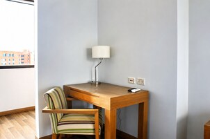 Quarto pequeno e bem arrumado com uma escrivaninha de madeira, uma cadeira listrada, um abajur e uma janela com vista para prédios.