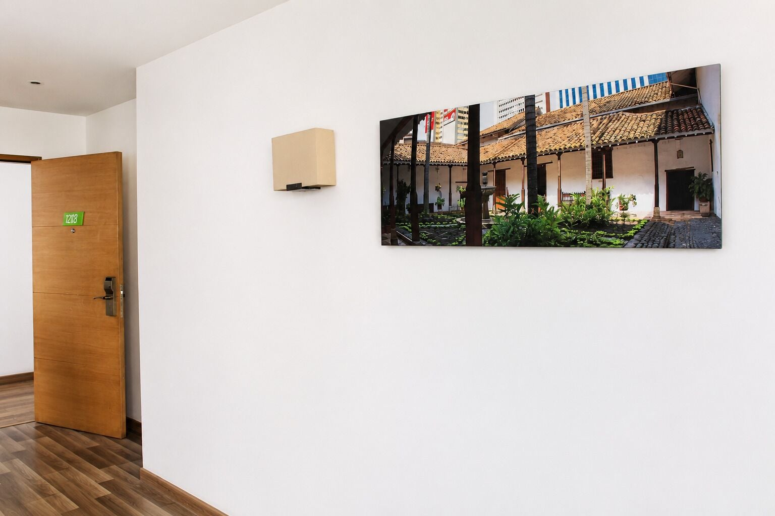 Um corredor com porta de madeira, luminária na parede e fotografia emoldurada de um pátio.