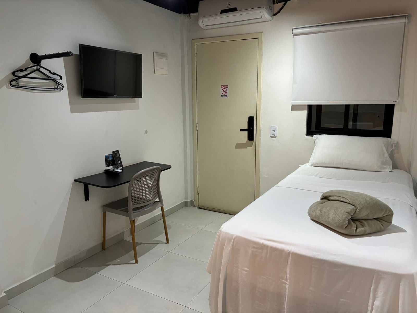 Quarto pequeno e limpo, com uma cama, uma escrivaninha, uma cadeira, uma TV e uma porta com o aviso 'não fumar'.