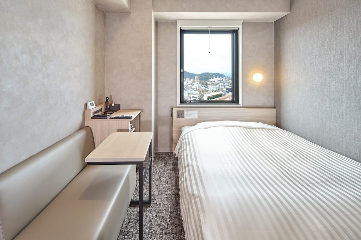 Quarto de hotel com cama, escrivaninha, cadeira, janela com vista para a cidade e luminária fixada na parede.