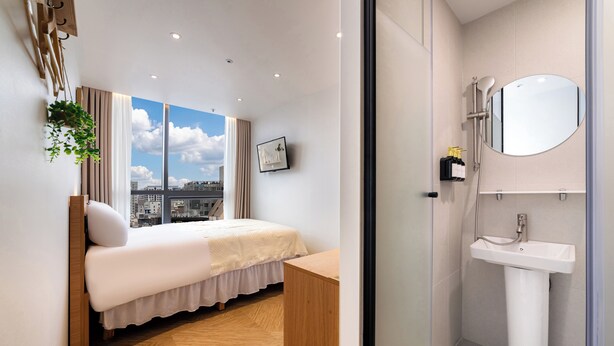 Quarto de hotel moderno com cama, mesa de cabeceira, grande janela com vista para a cidade, banheiro com pia e espelho, e um vaso de planta.