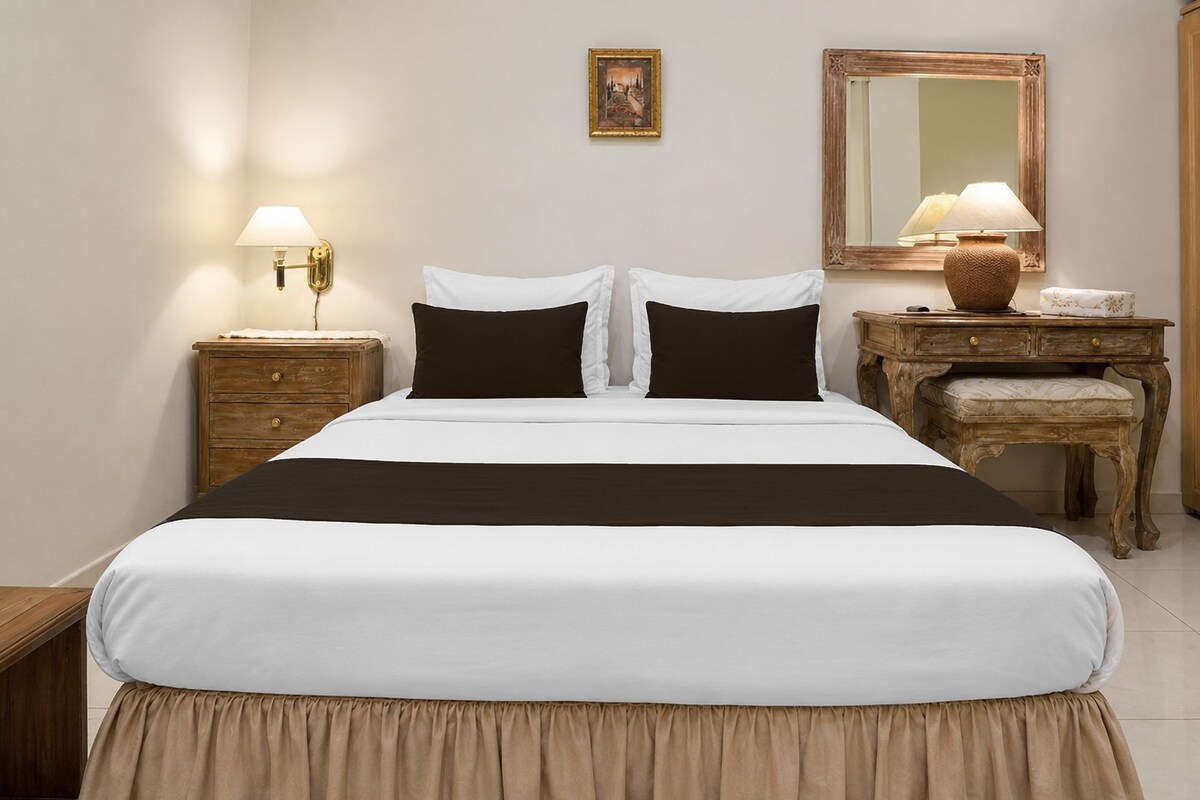 Cama bem arrumada com colcha branca e preta, duas mesas de cabeceira com abajures, um criado-mudo de madeira, um espelho e uma pequena mesa com um vaso.