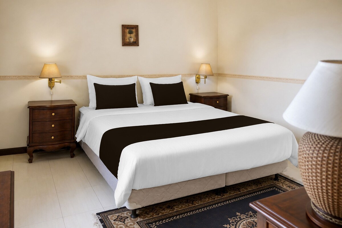 Quarto de hotel com uma cama grande, duas mesas de cabeceira, um criado-mudo, uma luminária e uma pequena moldura na parede.