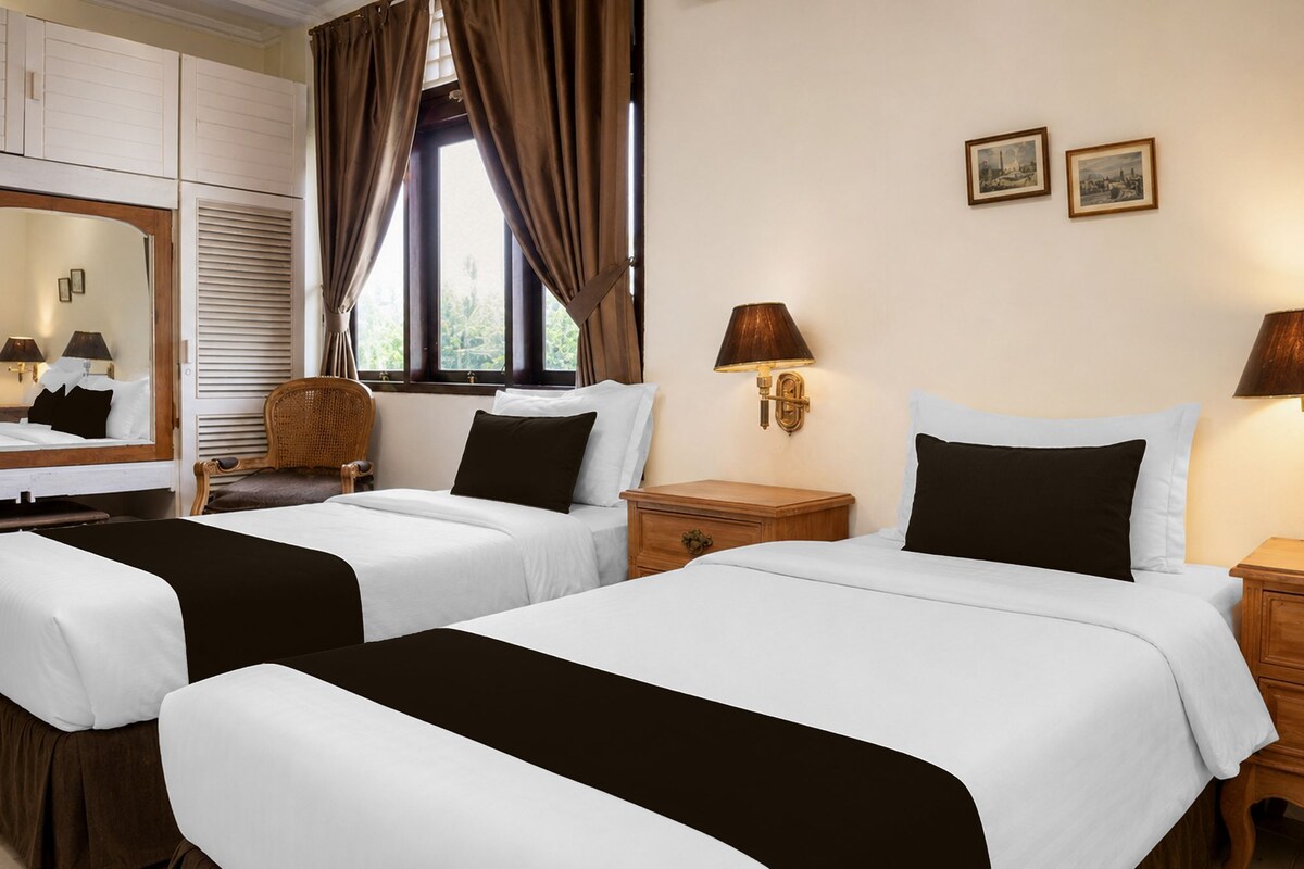 Quarto de hotel com duas camas, mesa de cabeceira de madeira, luminária, espelho e janela com cortinas.