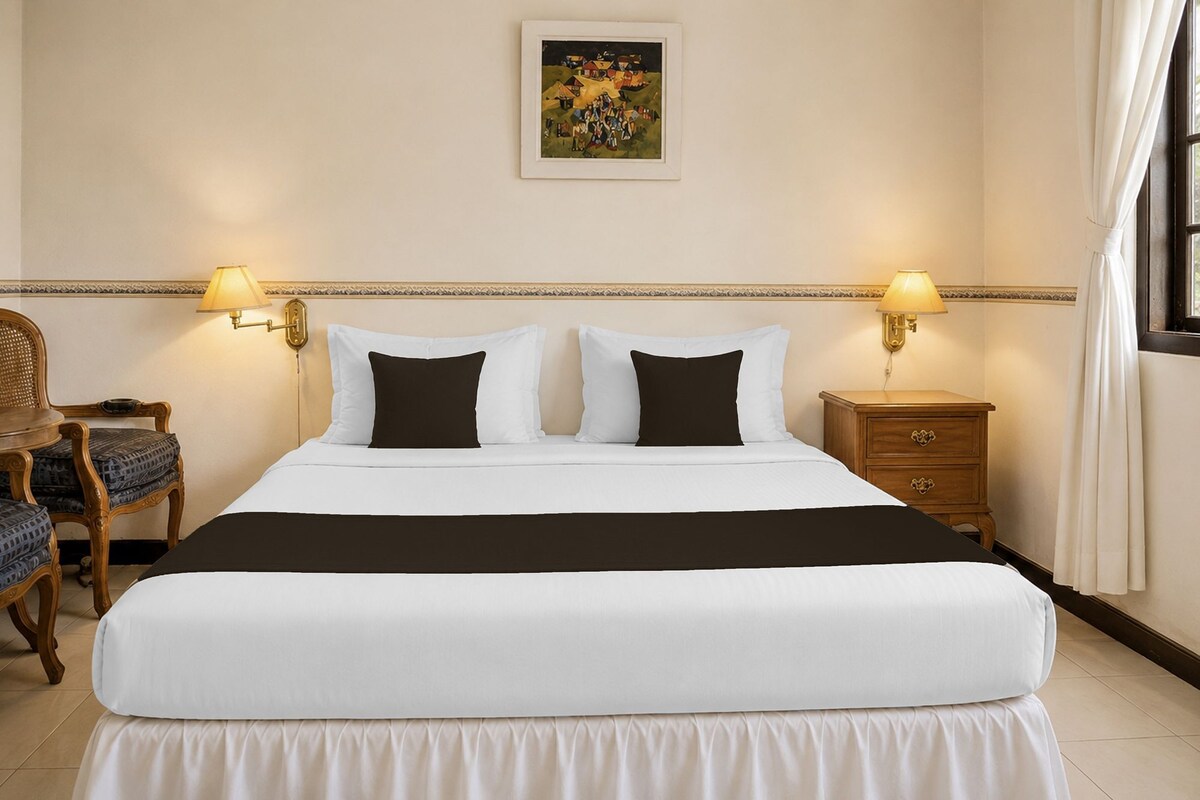 Quarto de hotel com cama, duas luminárias de cabeceira, mesa de apoio, uma cadeira e um quadro na parede.