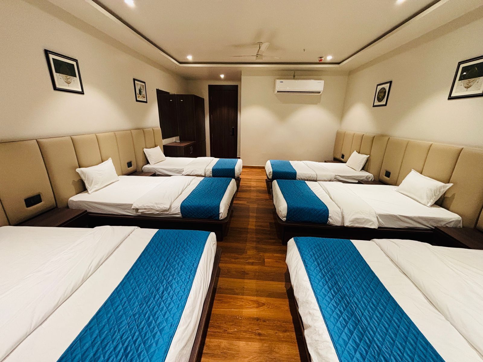 Quarto de hotel com quatro camas dispostas em linha, piso de madeira e um aparelho de ar condicionado na parede.