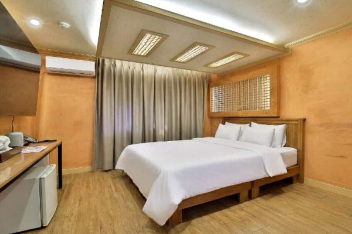Quarto de hotel com cama, escrivaninha com computador, aparelho de ar condicionado e janela com cortinas.