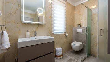 Casa de banho com parede de mármore, lavatório, sanita, cabine de duche em vidro e um porta-toalhas.