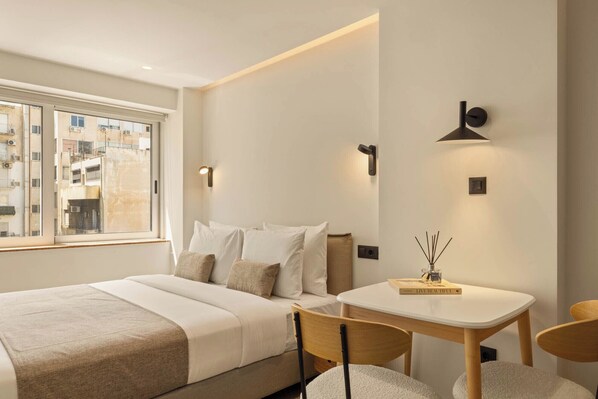 Quarto de hotel com cama, mesa pequena, duas cadeiras, janela e iluminação embutida na parede.