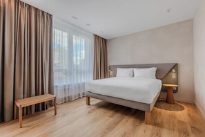 Quarto de hotel moderno com cama grande, um banco e vista da cidade através de cortinas translúcidas.