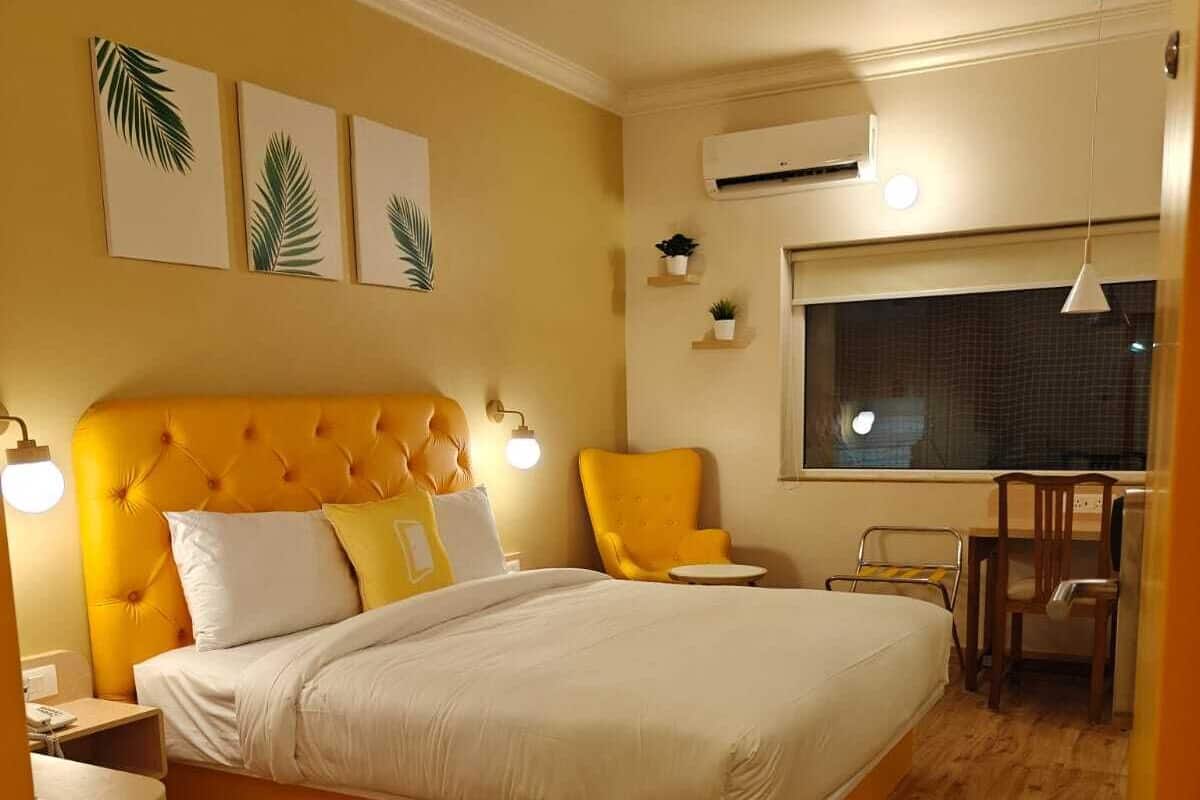 Quarto com uma cama grande, uma mesa de cabeceira, uma cadeira, uma pequena mesa e uma janela com ar condicionado.