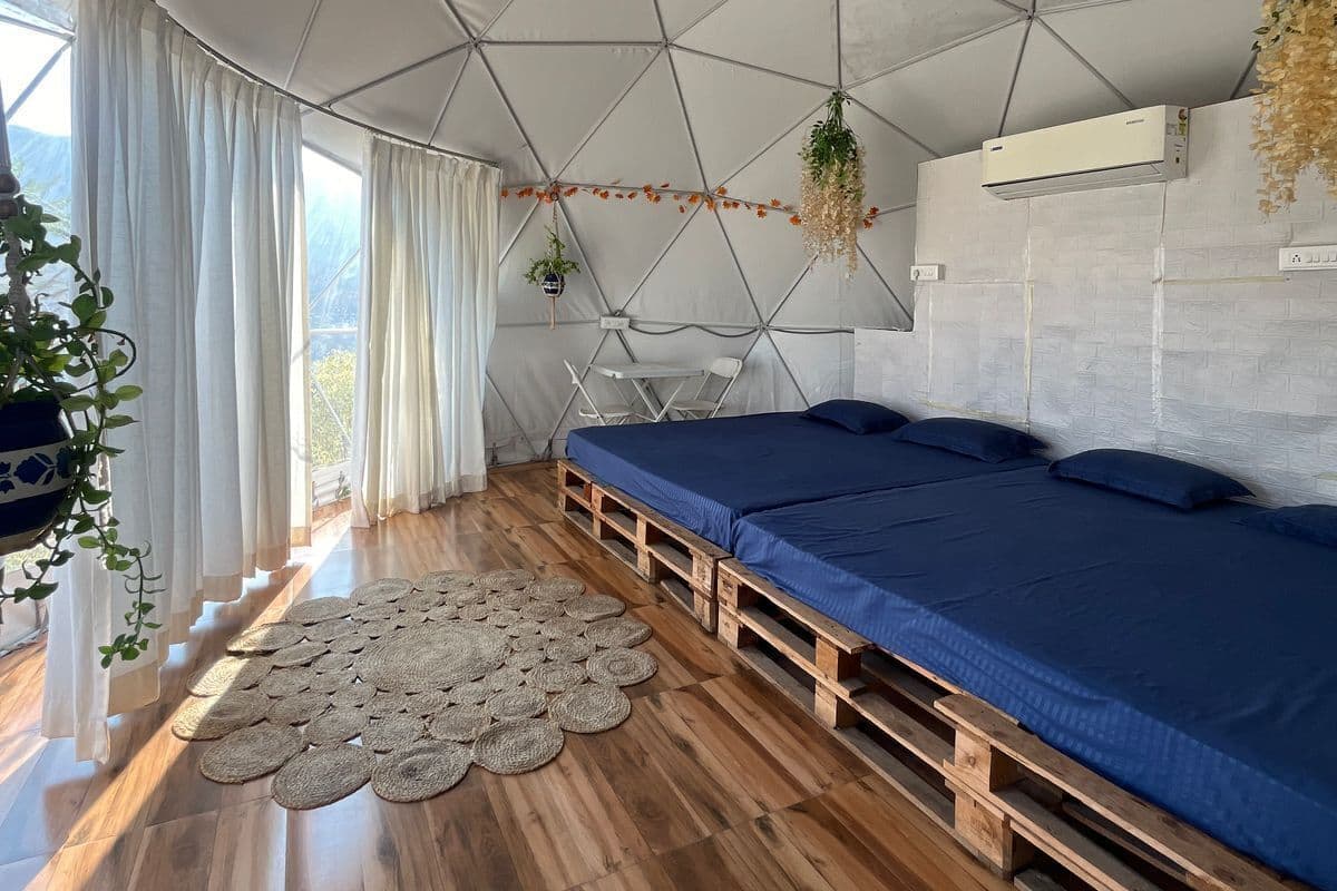 Uma tenda com piso de madeira, duas camas com roupa de cama azul, um candeeiro pendente e um ar condicionado fixado na parede.