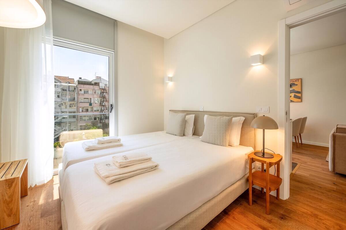 Quarto de hotel com uma cama grande, uma mesinha de cabeceira com abajur, um pequeno banco de madeira e vista da cidade pela janela.